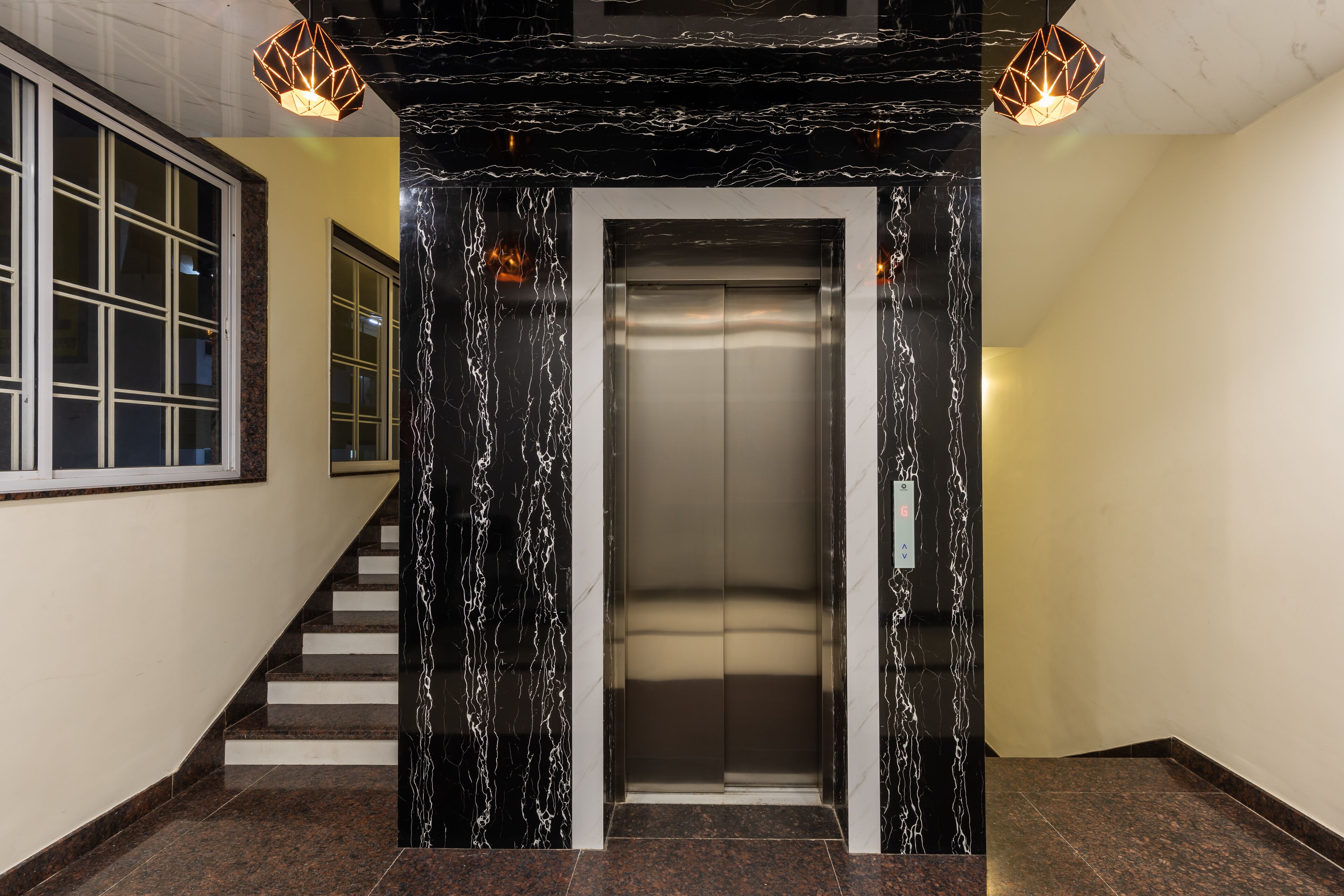 Um elevador com interior metálico, ladeado por paredes de mármore negro com veios brancos, e uma escadaria com degraus de mármore escuro.