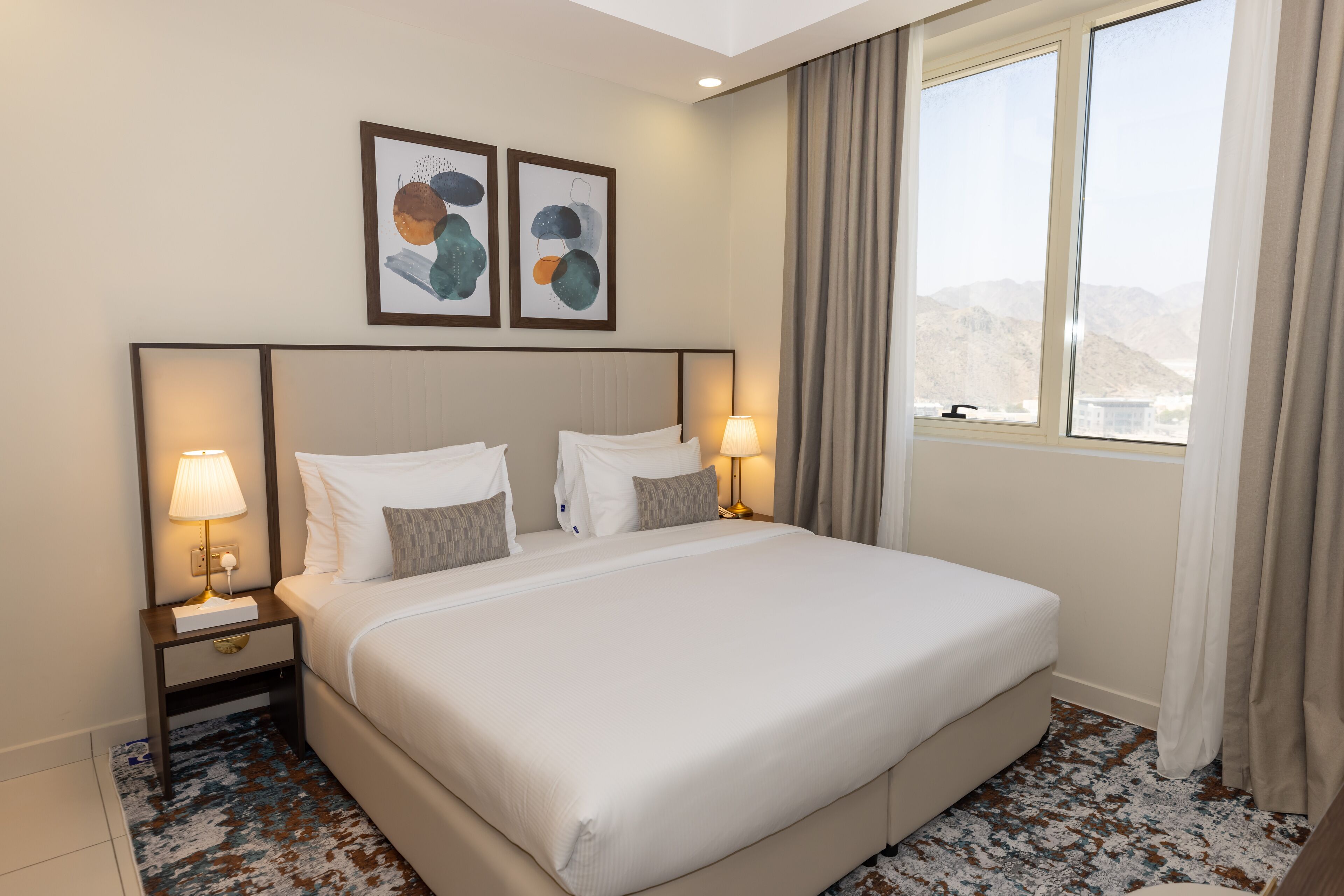 Quarto de hotel com uma cama grande, duas mesas de cabeceira com abajures, uma janela com cortinas e arte abstrata na parede.