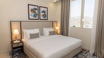 Quarto de hotel com uma cama grande, duas mesas de cabeceira com abajures, uma janela com cortinas e arte abstrata na parede.