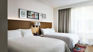 Quarto de hotel com duas camas, cabeceira, mesa de cabeceira, luminária e janela com cortinas.