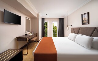 Quarto de hotel com cama, escrivaninha, cadeira, televisão, quadro emoldurado e janela com cortinas.