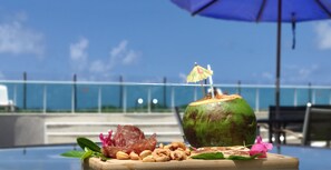 Disposição tropical com um coco, petiscos e um guarda-sol decorativo sobre uma mesa com vista para o mar.
