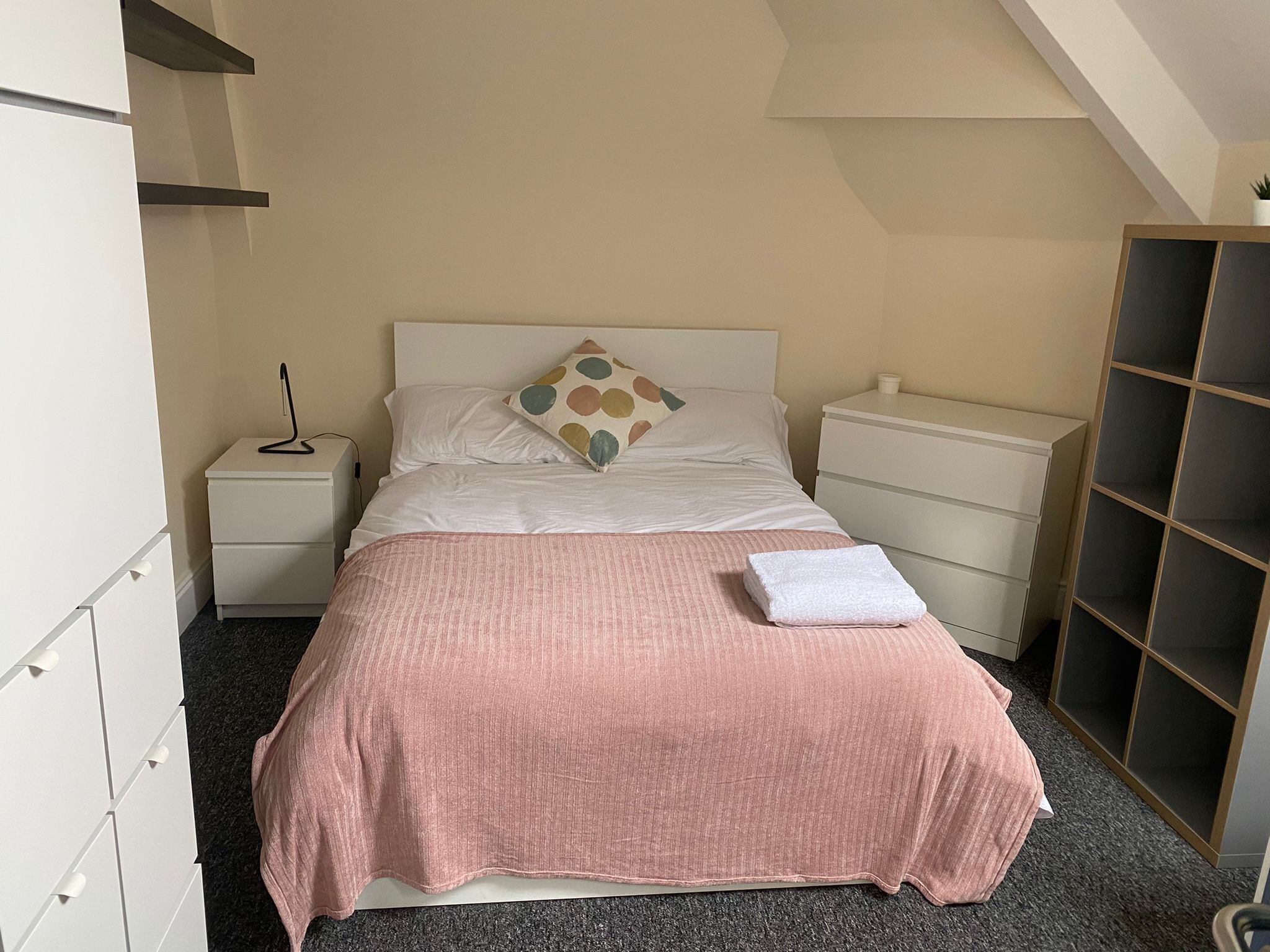 Cama bem arrumada com colcha e almofadas cor-de-rosa, mesa de cabeceira com abajur, criado-mudo e guarda-roupa com espelho.