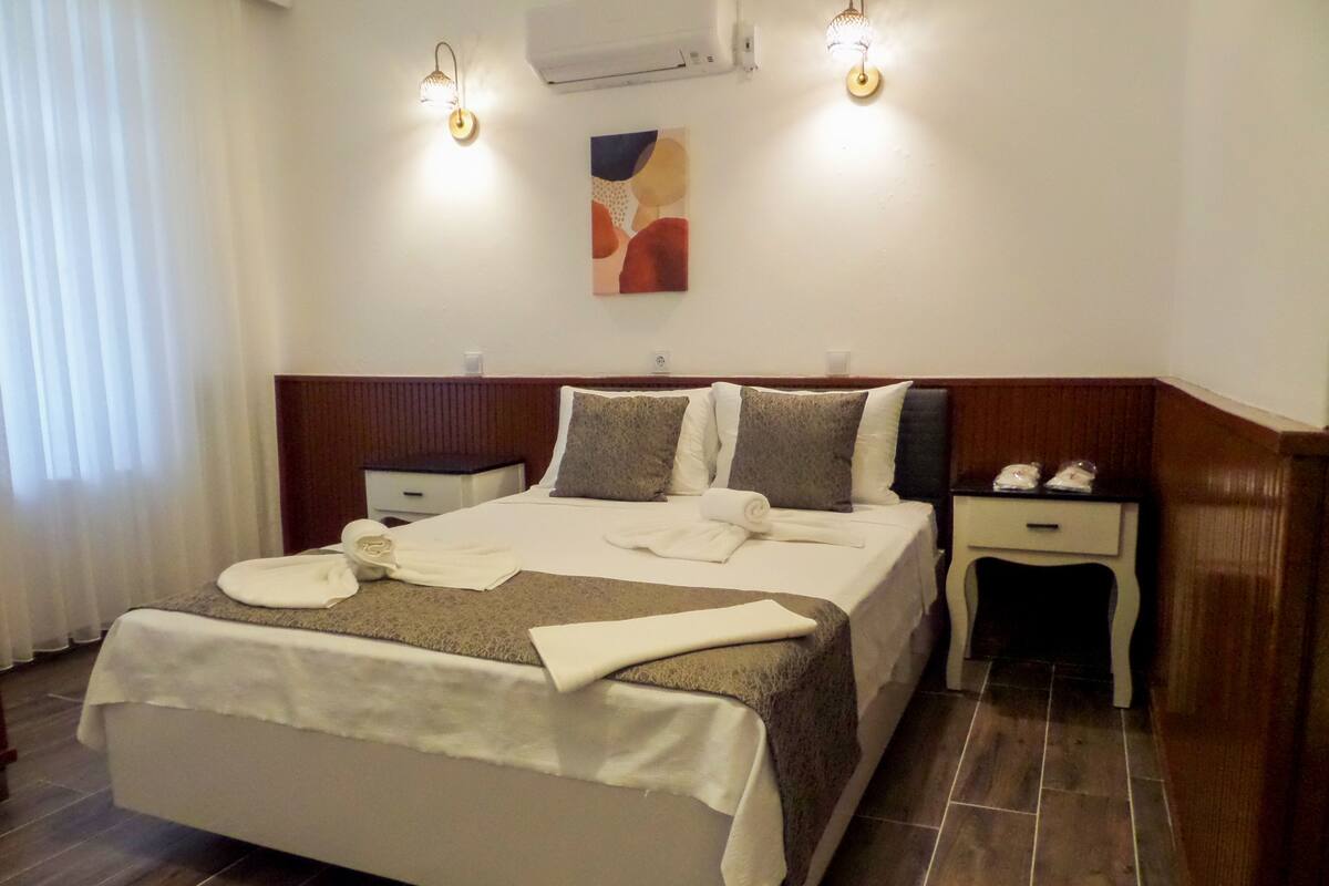 Uma cama bem arrumada com edredom branco e marrom, duas mesas de cabeceira com abajures, um ar condicionado fixado na parede e um quadro sobre a cama.