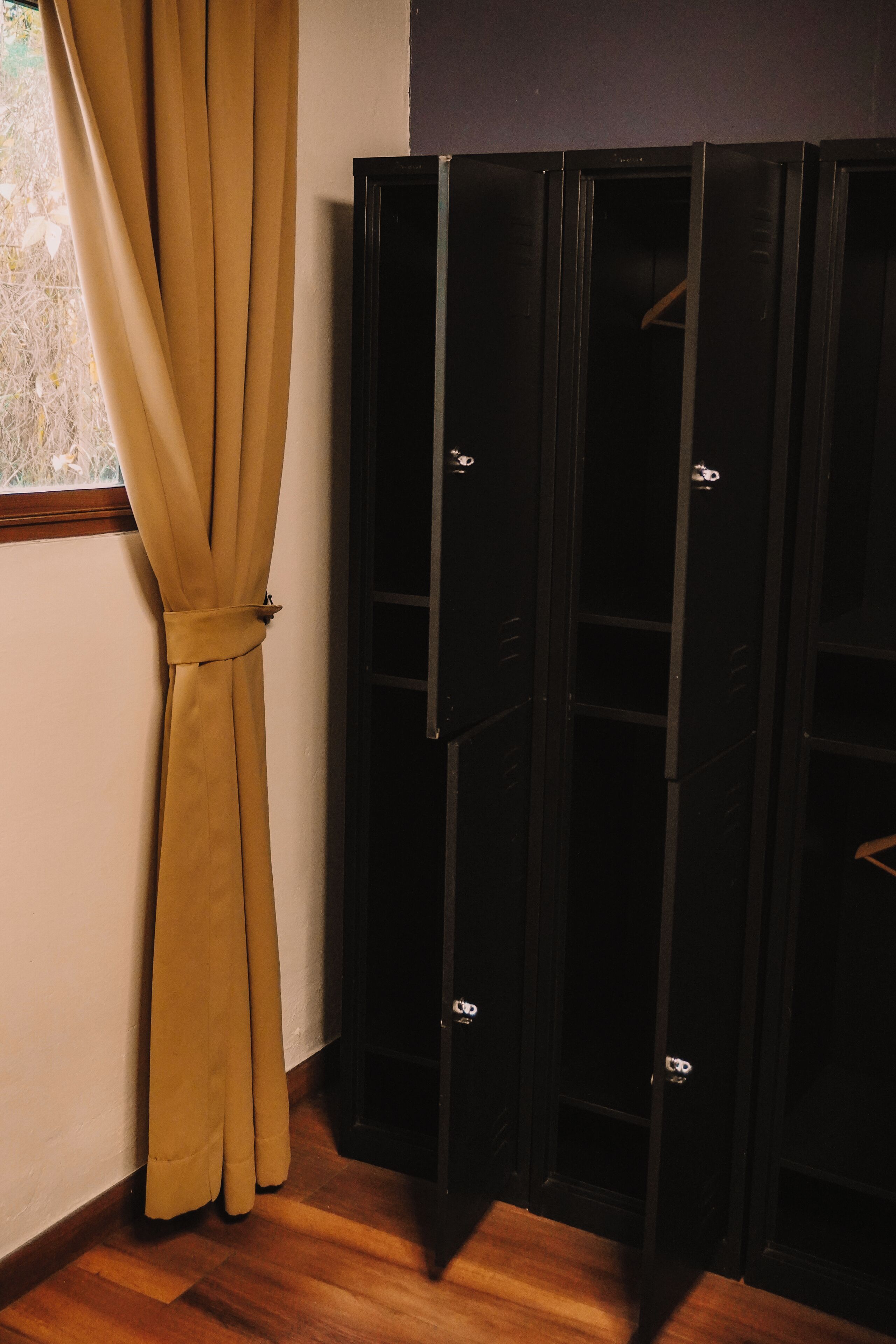 Quarto com piso de madeira, cortina bege e guarda-roupa preto com várias portas.