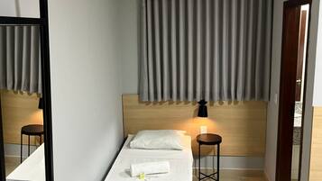 Quarto de hotel compacto com uma cama de solteiro, mesa de cabeceira com luminária, um banquinho pequeno e unidade de ar condicionado fixada na parede.