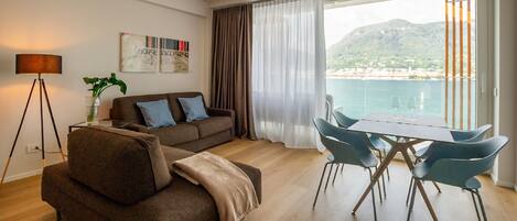 Uma sala de estar moderna com um sofá, uma mesa de jantar com cadeiras, um abajur e vista para o mar por grandes janelas.