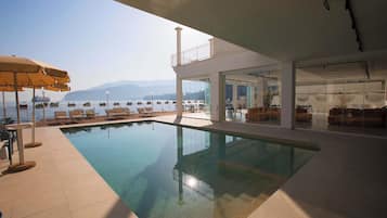 Uma área de piscina moderna com vista clara para o mar e para as montanhas, equipada com espreguiçadeiras e guarda-sóis.