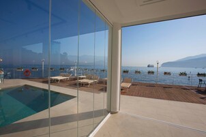 Uma área de piscina moderna com paredes de vidro transparente, cadeiras de lounge e vista para o mar.