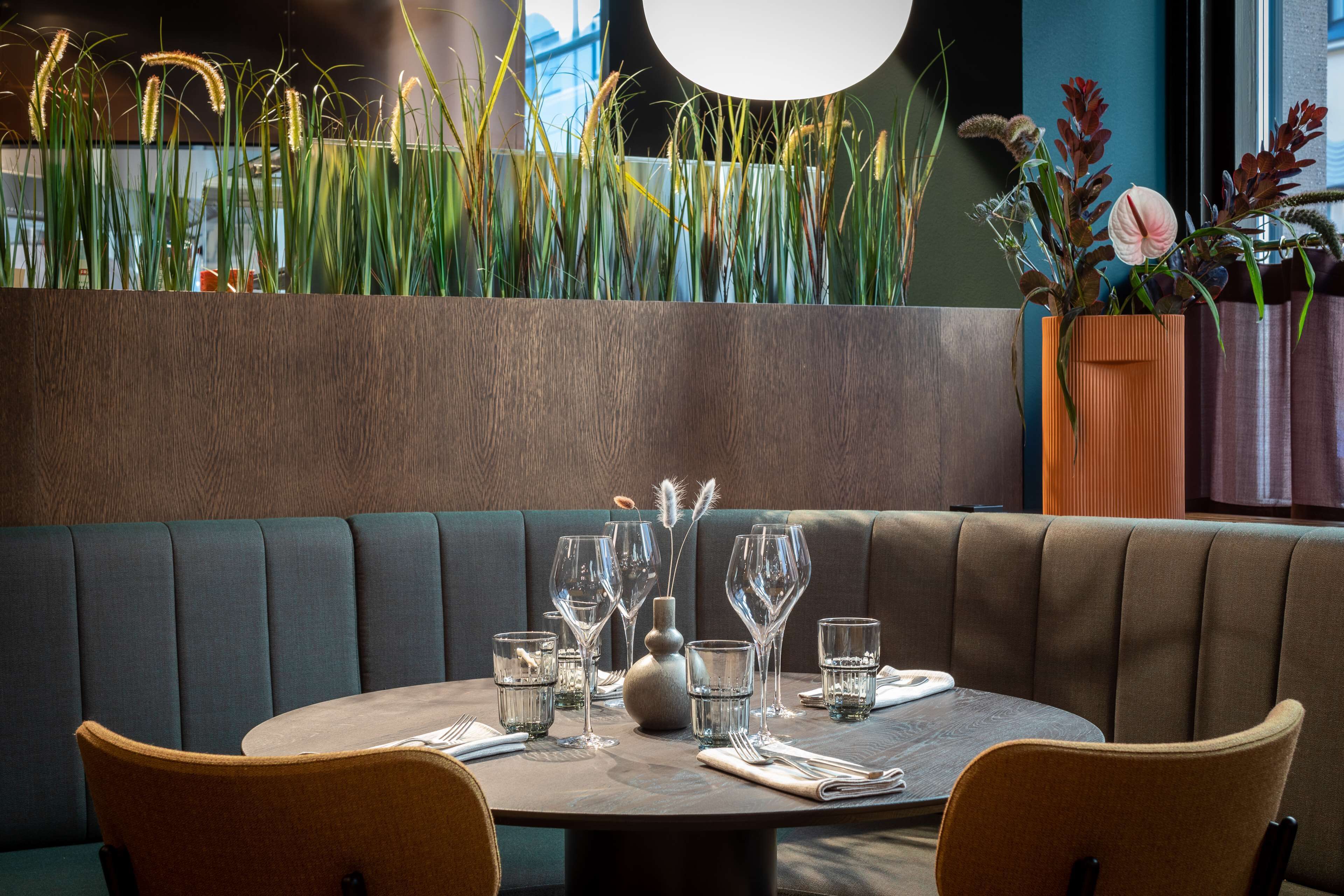 Um ambiente de restaurante com uma mesa redonda posta para refeições, um assento com banquinho estofado e uma parede decorativa com plantas.