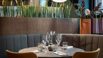 Um ambiente de restaurante com uma mesa redonda posta para refeições, um assento com banquinho estofado e uma parede decorativa com plantas.