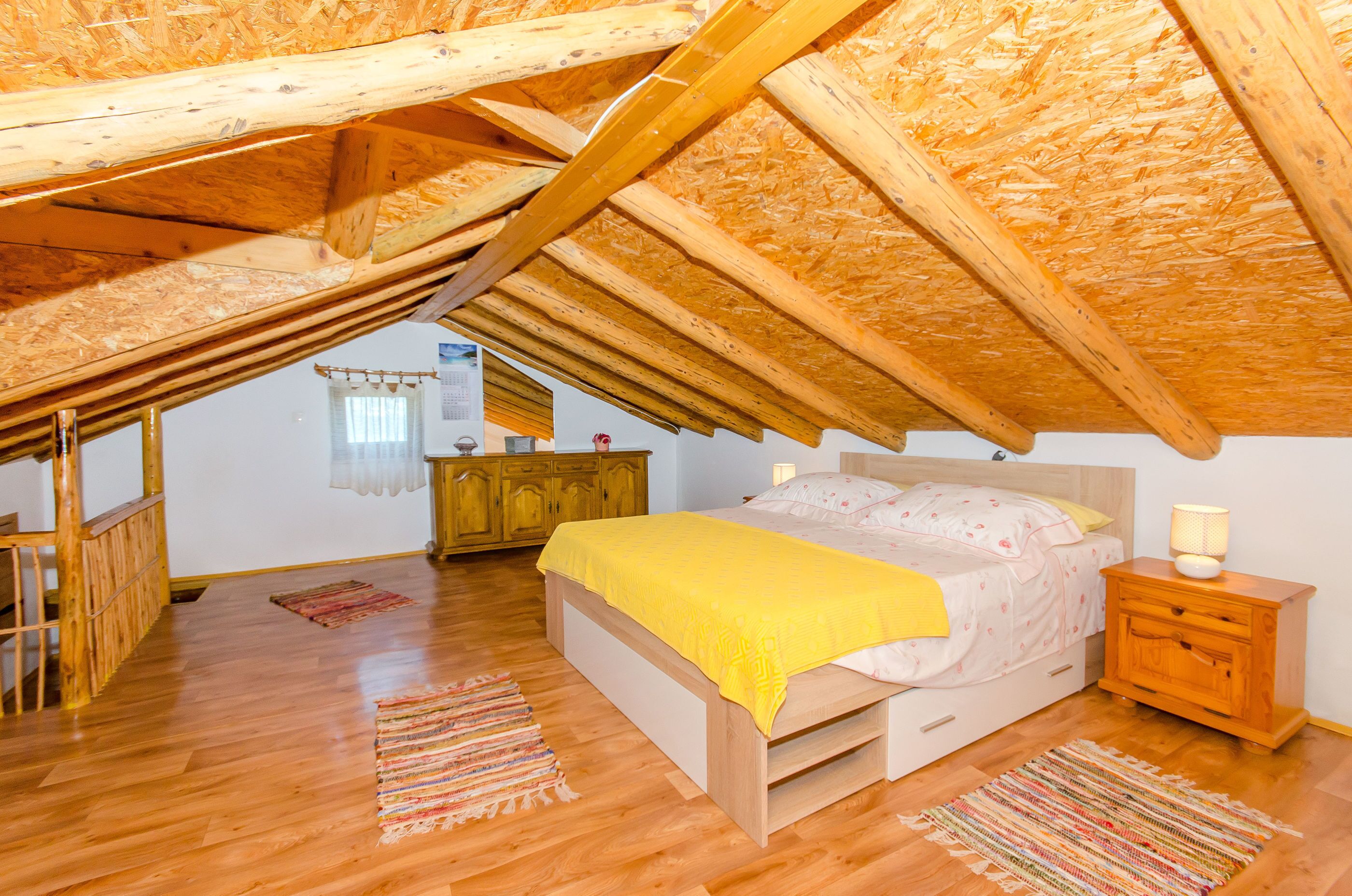 Quarto com teto de madeira inclinado, cama com manta amarela, mesinhas de cabeceira de madeira e cómoda de madeira.