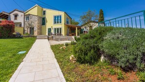 Uma casa amarela de dois andares com fachada de pedra, um caminho pavimentado que leva à entrada e uma cerca verde ao fundo.