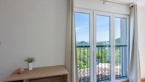 Quarto com uma escrivaninha de madeira, uma pequena planta em vaso e vista para uma varanda com grades.
