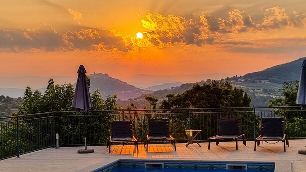 Área de piscina com espreguiçadeiras e guarda-sóis, com vista para uma paisagem deslumbrante com colinas e um pôr do sol.