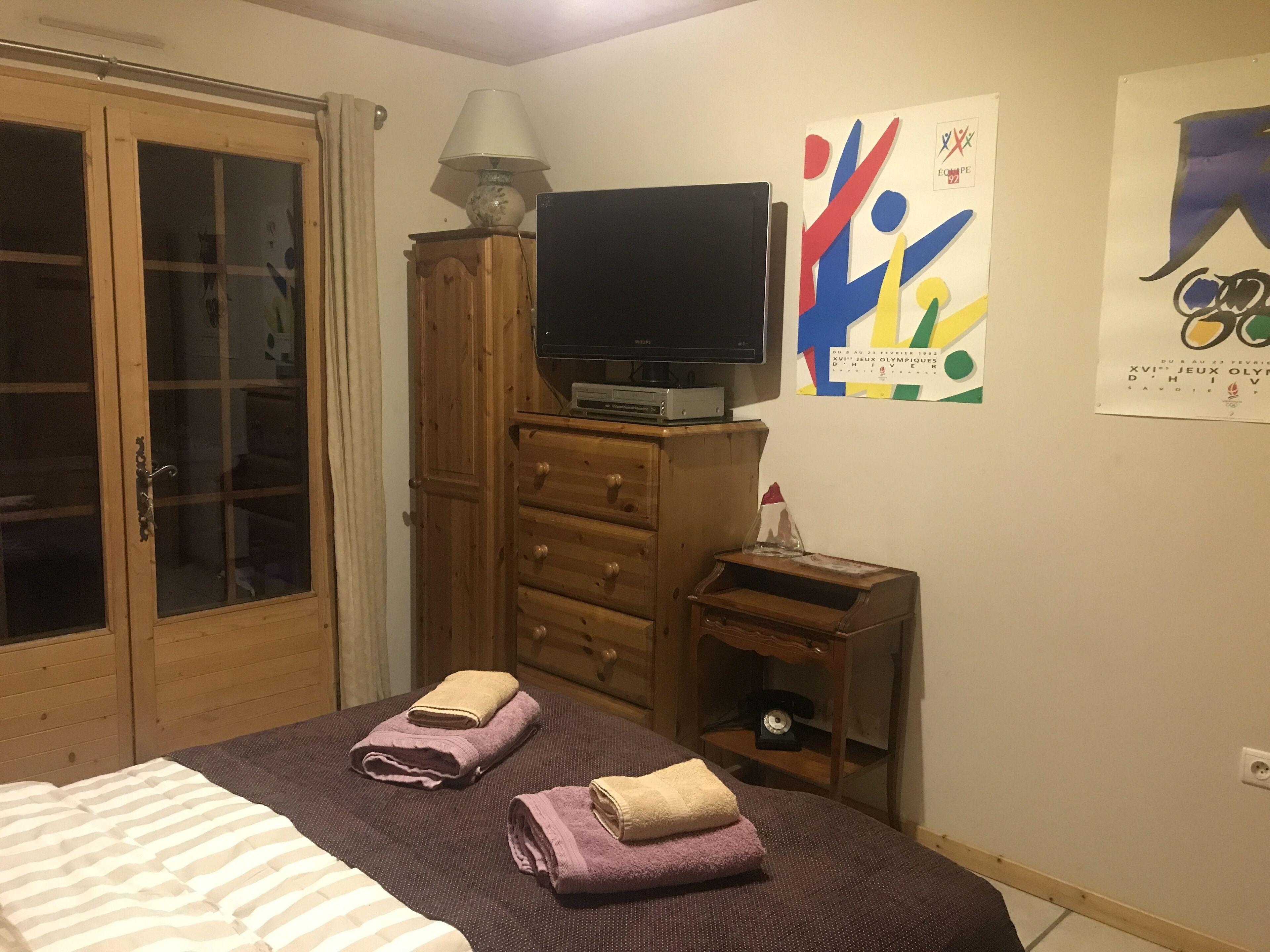 Quarto com cama, guarda-roupa de madeira, televisão em cima e uma pequena mesa com abajur.