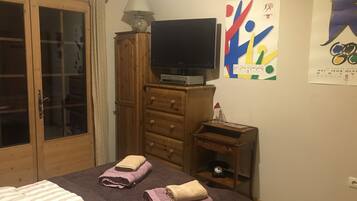 Quarto com cama, guarda-roupa de madeira, televisão em cima e uma pequena mesa com abajur.