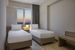 Quarto de hotel com duas camas, um banco, janela com cortinas e vista para a paisagem urbana ao entardecer.