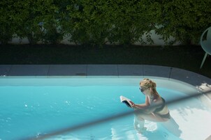 Alguém lendo um livro perto de uma piscina, com uma sebe verde ao fundo.