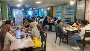 Restaurante com várias mesas e cadeiras, pessoas jantando e vista da cidade por grandes janelas.