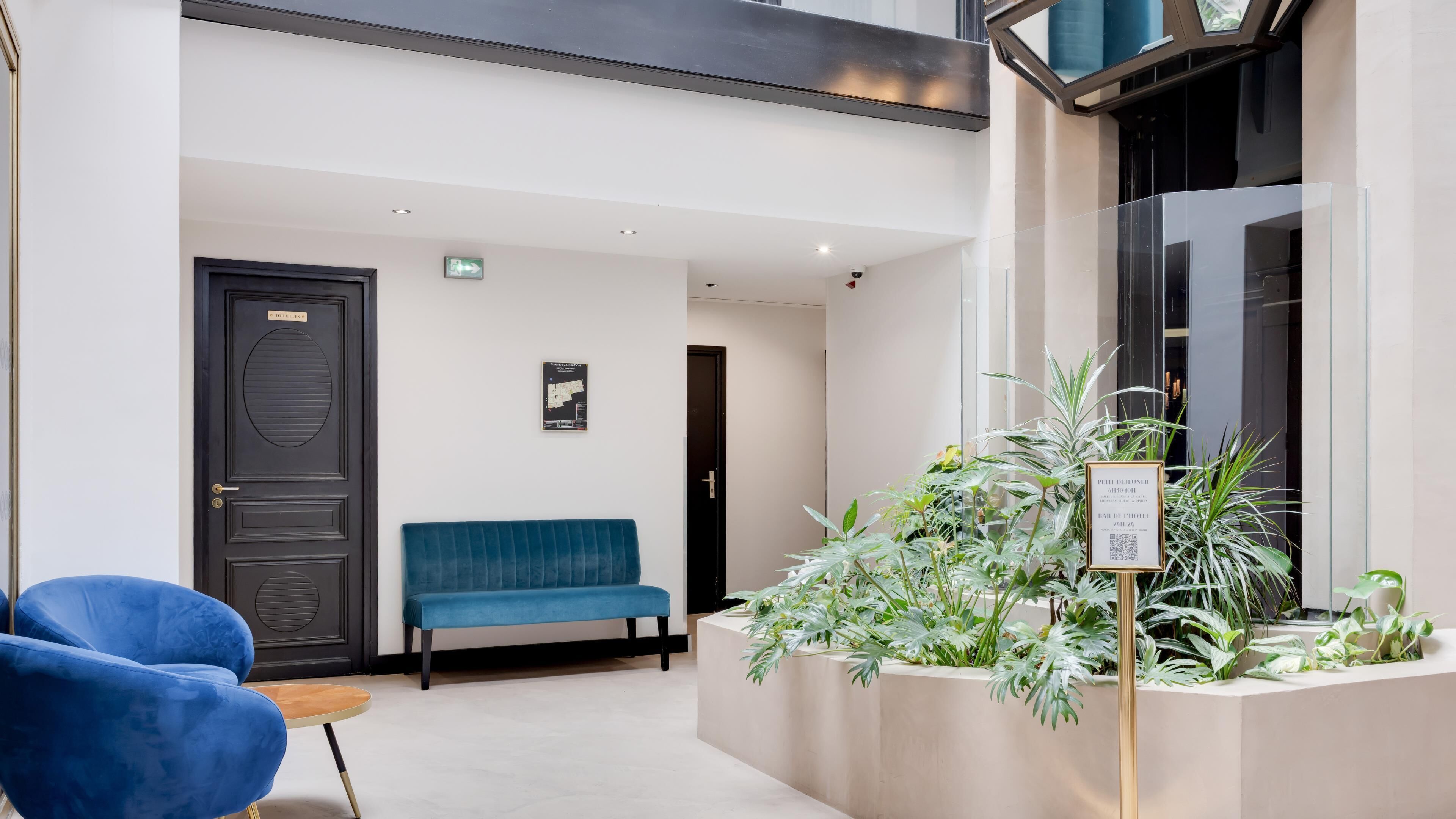 Um hall de hotel moderno com um sofá azul, uma mesa redonda pequena e um arranjo de plantas em vaso.