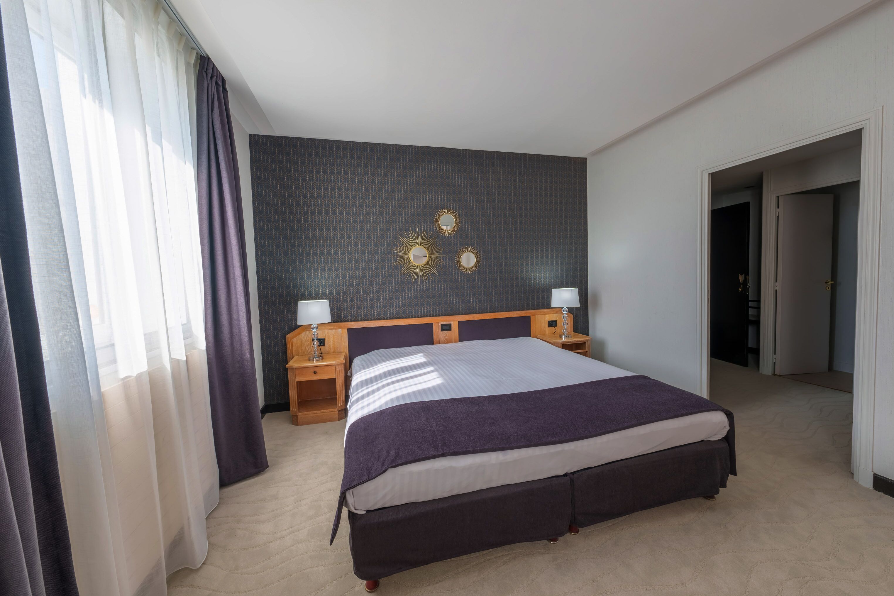 Quarto de hotel com uma cama grande, duas mesas de cabeceira, um relógio de parede e uma porta que dá para outro quarto.