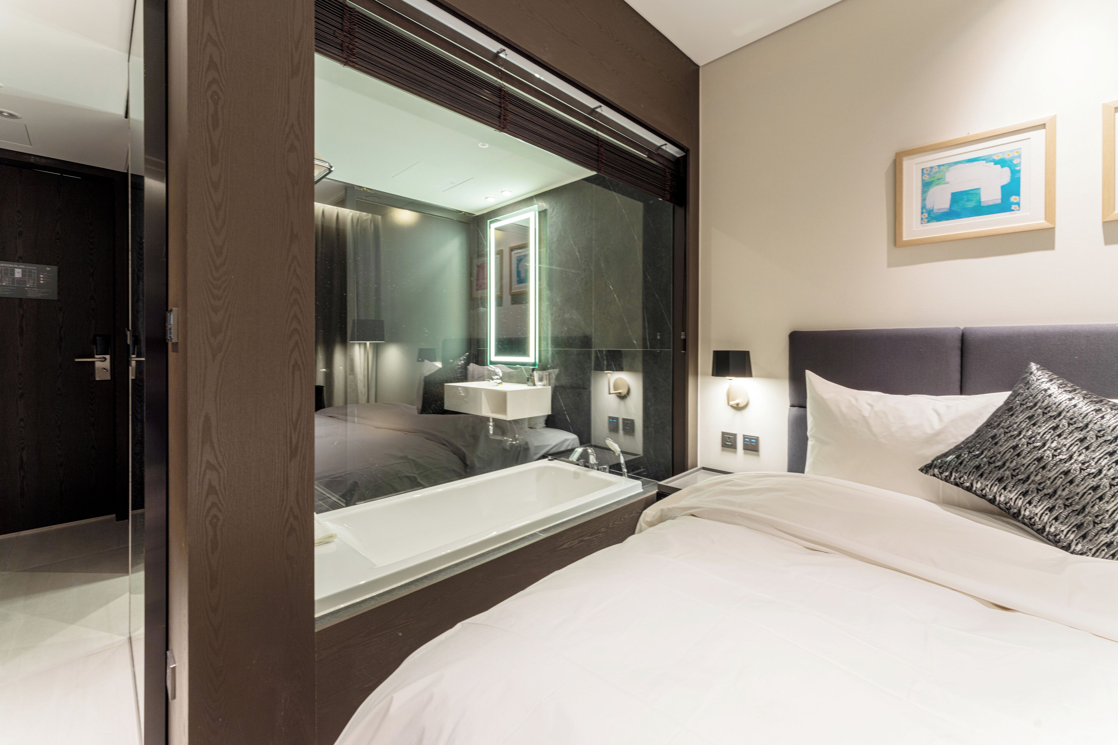 Quarto de hotel moderno com um espelho grande, uma banheira, uma cama com roupa de cama branca e duas fotos emolduradas na parede.