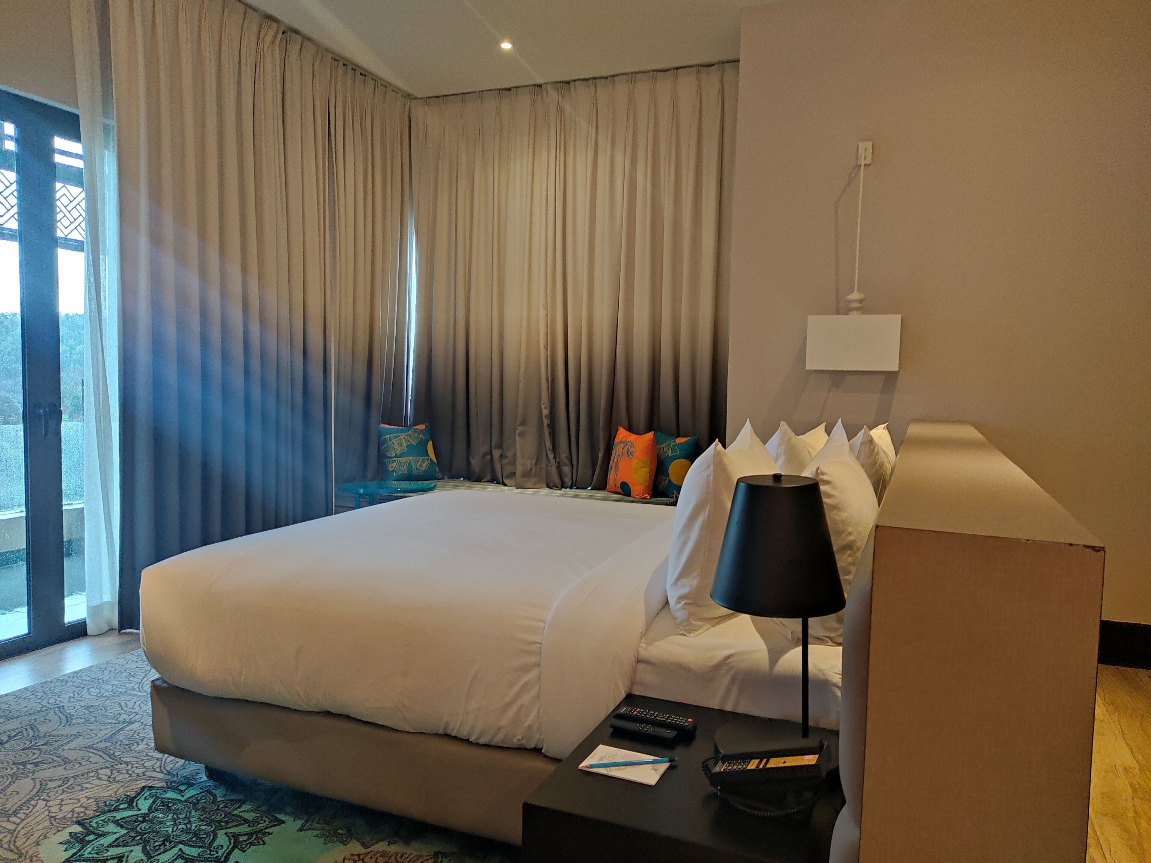 Quarto de hotel com uma cama grande, uma mesa de cabeceira com abajur e vista para o exterior através de uma janela com cortinas.