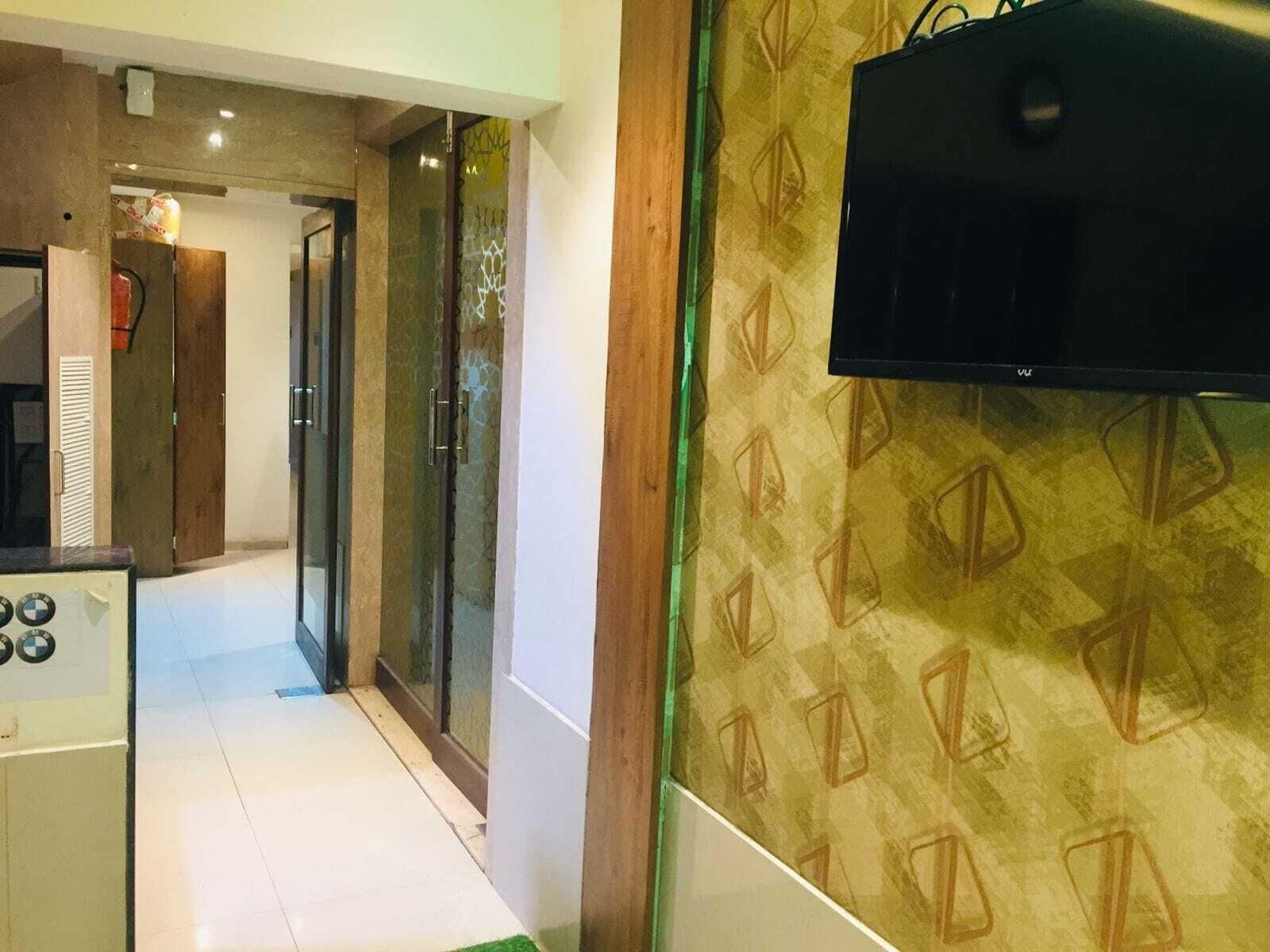 Um corredor de hotel com design moderno, apresentando uma porta de vidro, uma televisão de tela plana na parede e um painel de controle à esquerda.