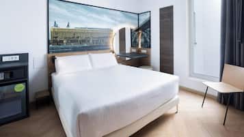 Quarto de hotel moderno com uma cama grande, uma escrivaninha com computador, uma cadeira e uma janela ampla.