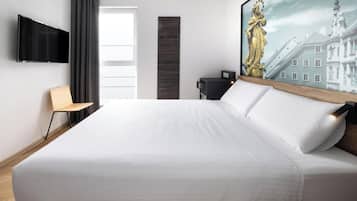 Quarto de hotel moderno com uma cama grande, televisão de tela plana na parede, escrivaninha com cadeira e um quadro de um edifício.