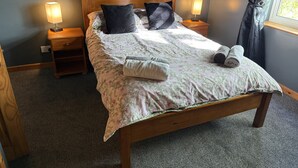 Uma cama bem arrumada com um cobre-leito floral, duas almofadas e toalhas dobradas sobre uma estrutura de madeira, num quarto com paredes escuras e uma janela com cortinas.
