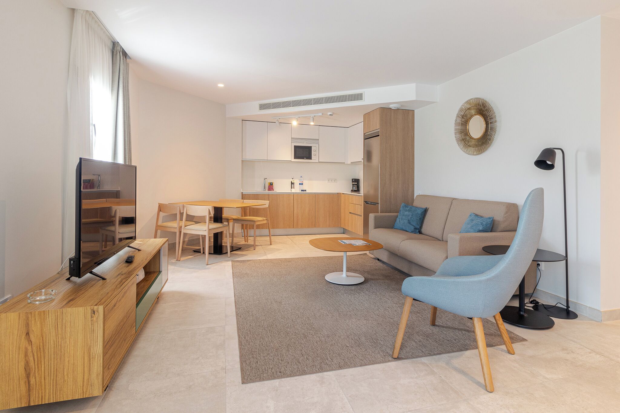 Uma sala de estar moderna com sofá, mesa de centro e painel para TV. Há uma cozinha com micro-ondas e geladeira.