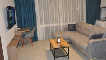 Uma sala de estar moderna com um sofá cinzento, uma mesa de centro de madeira, uma escrivaninha com uma cadeira e uma janela grande com cortinas leves.