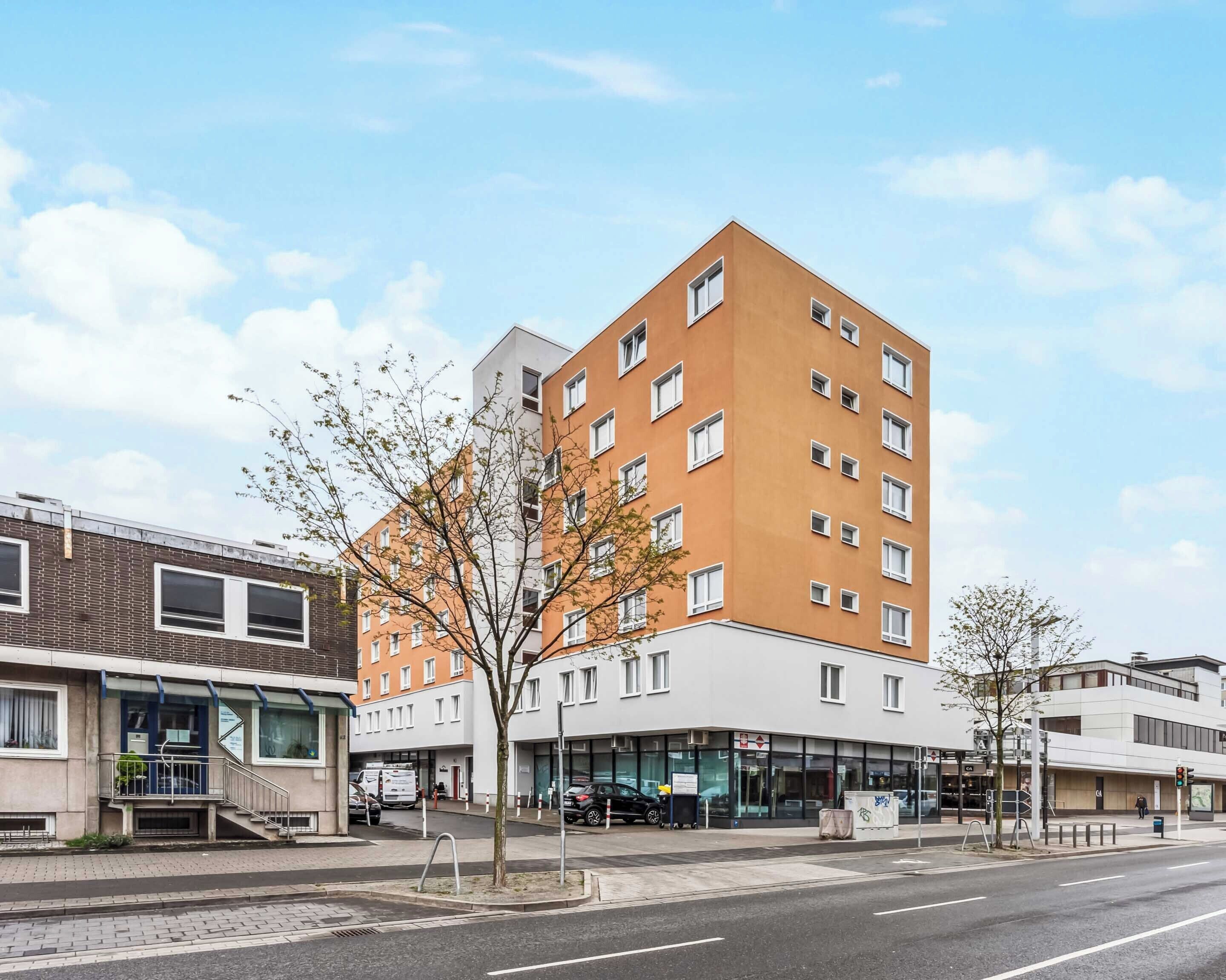 Um edifício moderno de vários andares com espaços comerciais e residenciais, destacando-se pela fachada laranja vibrante, grandes janelas e céu claro.