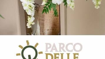Um corredor com um arranjo floral branco decorativo e o logotipo de "PARCO DELLE QUERCE" com um símbolo de sol e quatro estrelas.