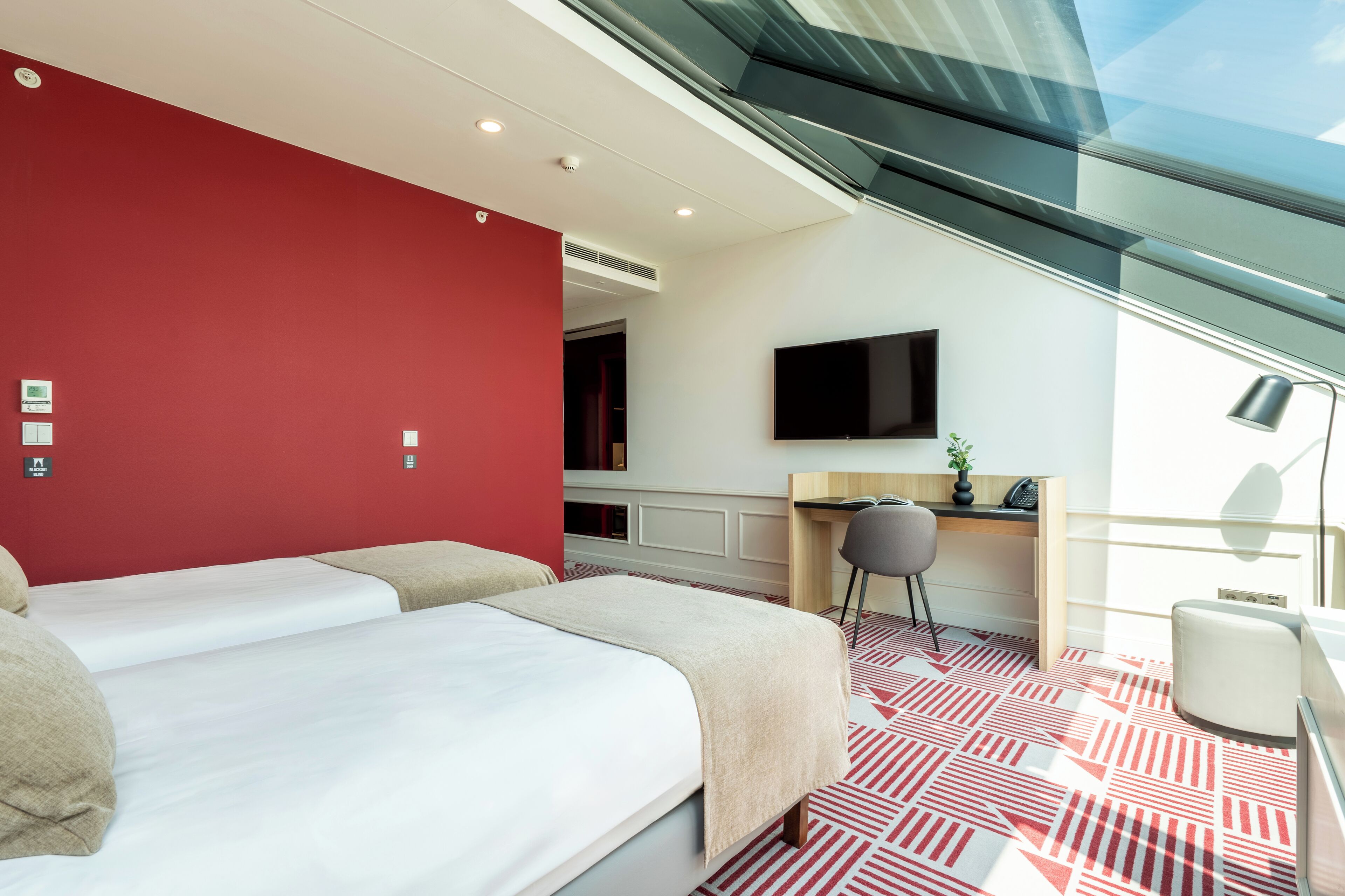 Quarto de hotel moderno com uma parede de destaque vermelha, uma cama, uma escrivaninha com cadeira, uma televisão e uma janela grande.