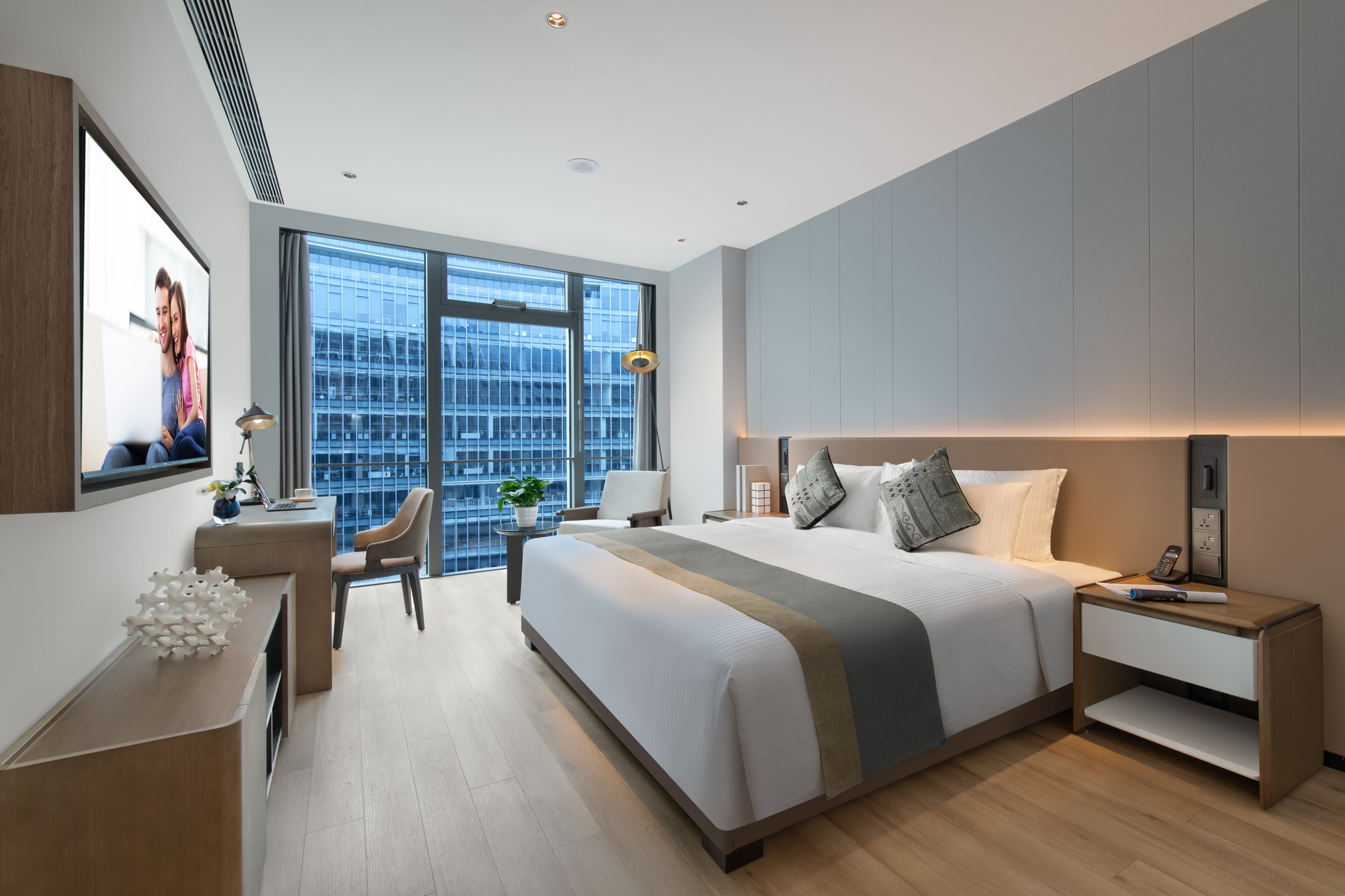 Quarto de hotel moderno com uma cama grande, uma escrivaninha, uma cadeira e vista para a cidade através de amplas janelas.