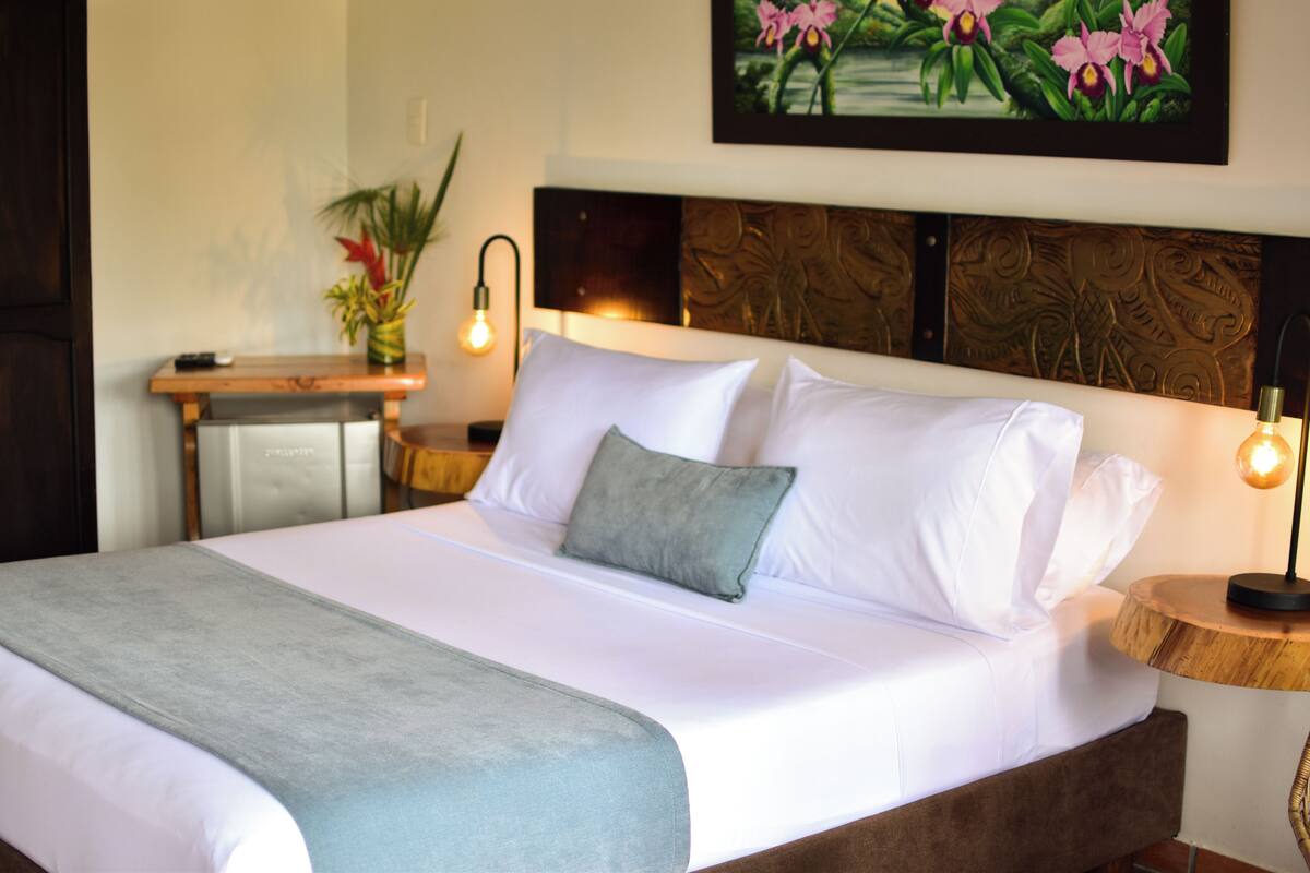 Cama bem arrumada com lençóis brancos e manta cinza, mesa de cabeceira de madeira com abajur e pintura de flores na parede.