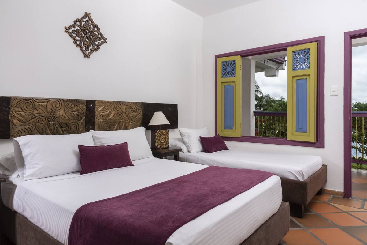 Quarto de hotel com duas camas, uma cabeceira decorativa, mesa de cabeceira e janela com vista para o verde.