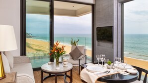 Quarto moderno com varanda e vista para a praia, mobiliado com sofá, mesa de jantar posta para dois e TV na parede.