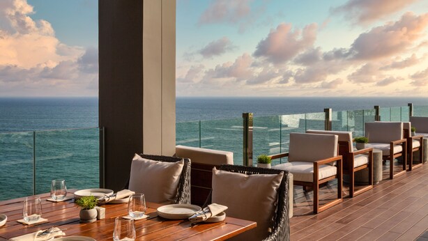 Uma área de refeições com vista para o oceano, com uma mesa de madeira, cadeiras e guarda-corpo de vidro.