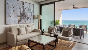 Uma sala de estar moderna com sofá, mesa de centro e cadeiras, um grande quadro abstrato e vista para o oceano através de grandes portas de vidro.