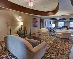 Um saguão de hotel com carpete estampado, sofás, mesa de centro, um vaso com flores, uma mesa com abajur e uma parede com quadros.
