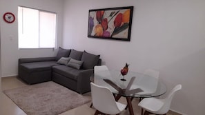 Uma sala de estar com sofá seccional cinza, mesa de centro de vidro e cadeiras brancas. Há um quadro na parede e um relógio na parede.
