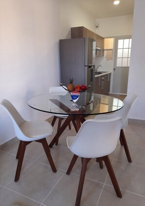 Uma sala de jantar com mesa de vidro rodeada por cadeiras brancas, um frigorífico e uma cozinha ao fundo.