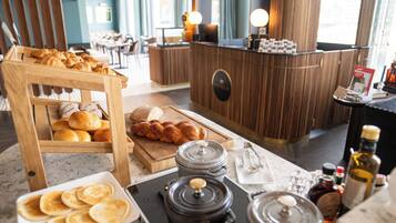 Um buffet de café da manhã com pães, bolos e panquecas, um balcão de recepção de madeira e uma área de bar com garrafas e copos.
