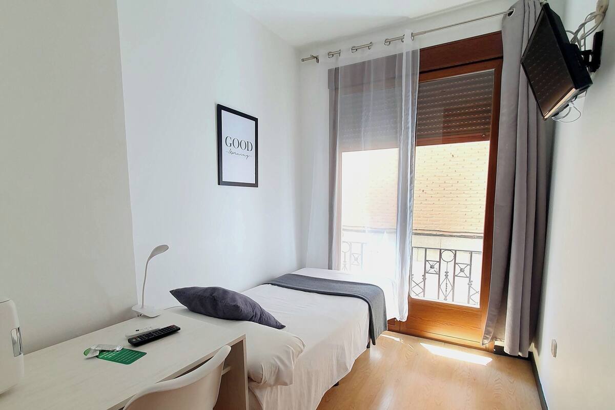 Quarto pequeno e limpo com uma cama, uma mesa e uma cadeira. Há uma janela com cortinas e vista para uma varanda.