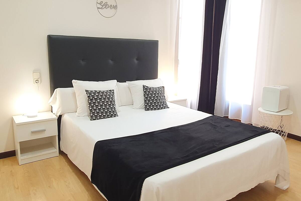 Cama bem arrumada com colcha preta e branca, duas almofadas iguais, mesa de cabeceira com abajur e luminária na parede.