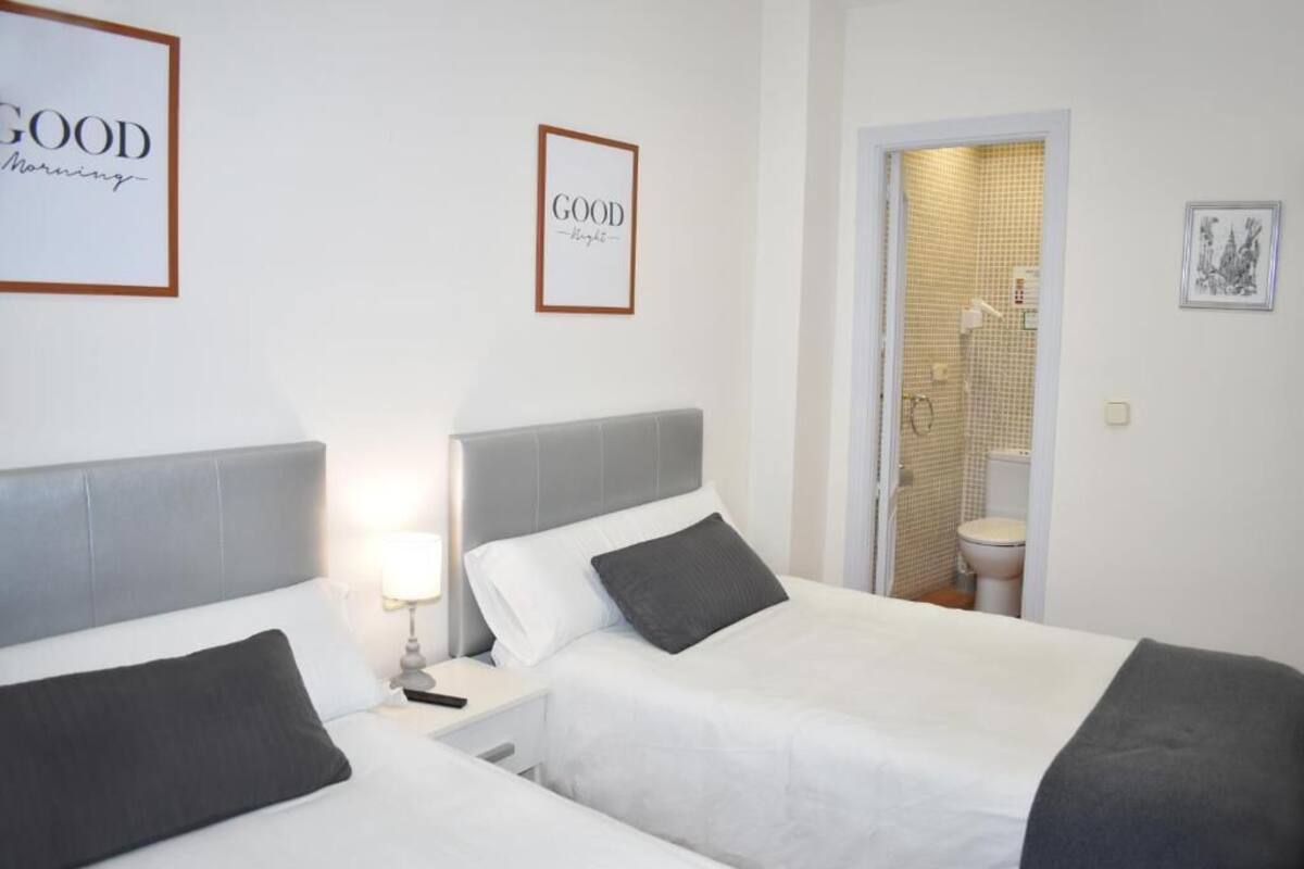Quarto de hotel com duas camas, uma mesa de cabeceira com abajur e banheiro visível por uma porta aberta.